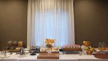 Disposição de buffet de café da manhã com diversos alimentos, incluindo pães, doces e frutas, sobre uma mesa com toalha branca.