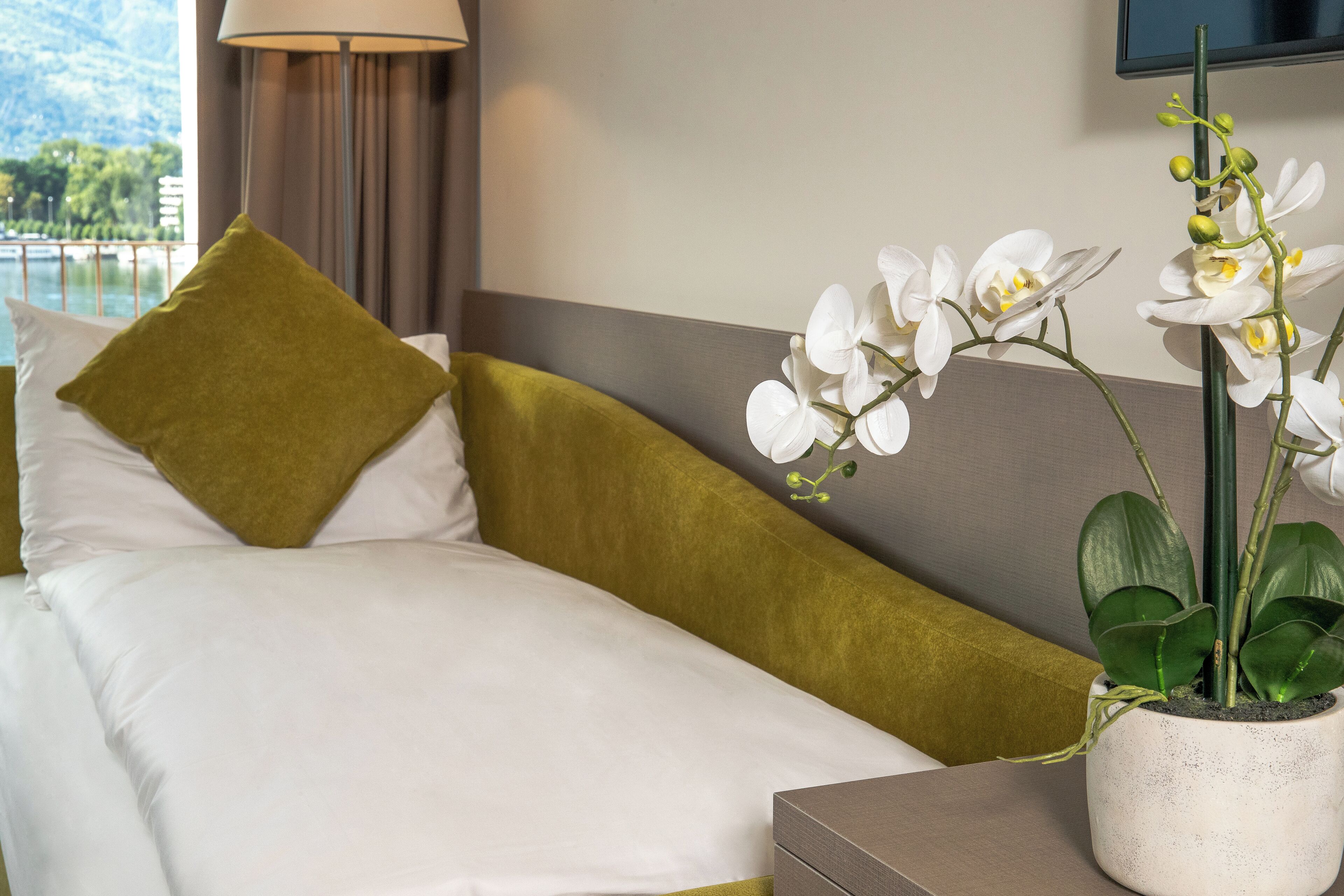 Quarto de hotel com uma cama, um travesseiro verde de destaque, um edredom branco e uma orquídea em vaso sobre uma mesa de cabeceira.