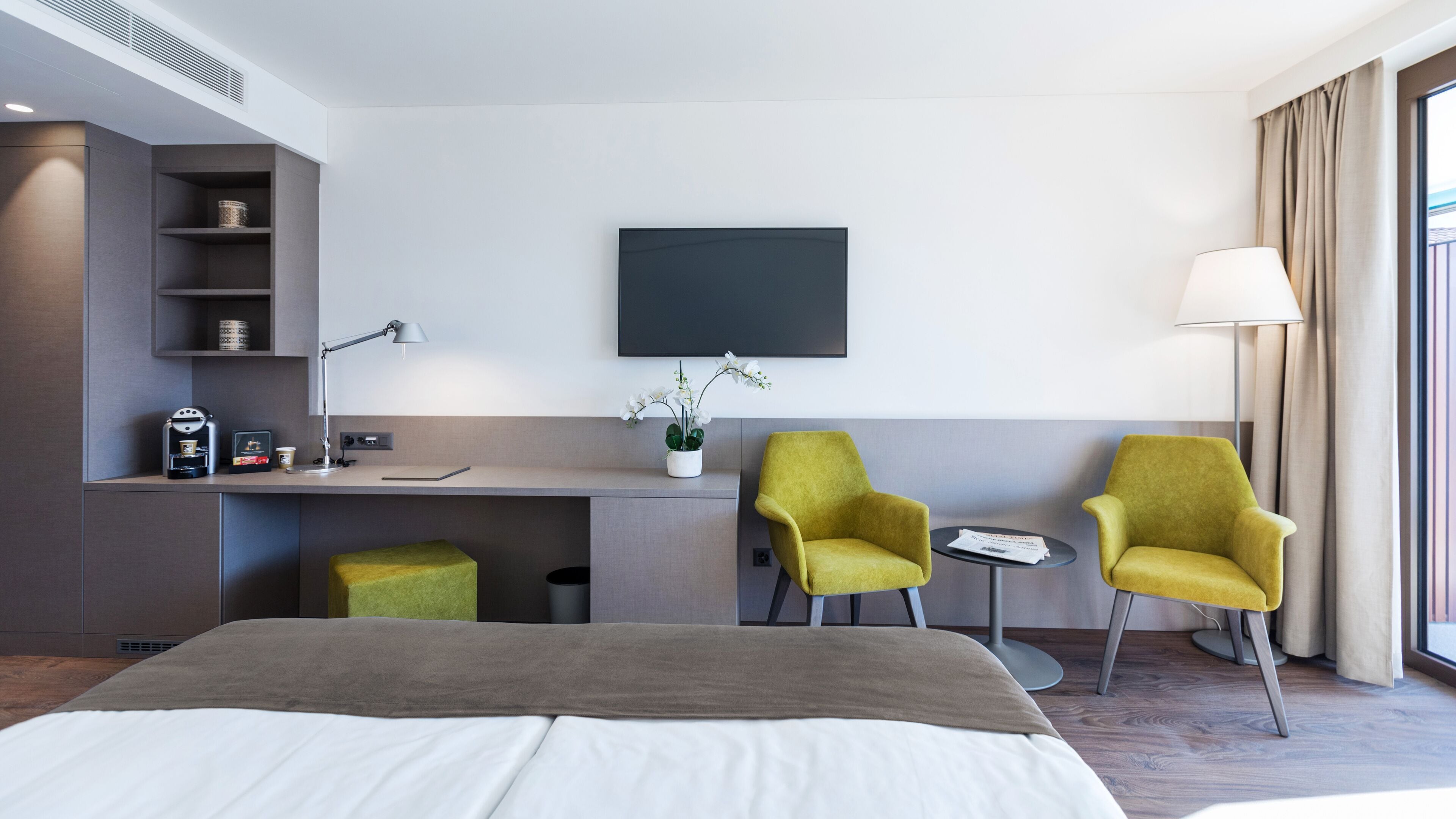 Quarto de hotel com cama, escrivaninha com cadeira, televisão, luminária e janela com cortinas.