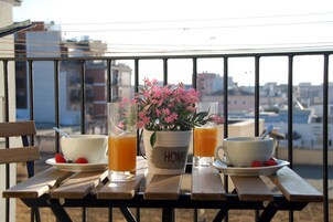 Varanda com uma mesa posta para o café da manhã, incluindo potes de iogurte e suco de laranja, uma planta em vaso e uma placa com a palavra 'HOME'.
