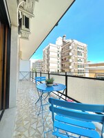 Varanda com cadeiras azuis e uma mesa pequena, chão de azulejos e vista para edifícios ao fundo.