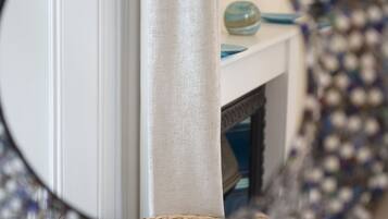 Um espelho com moldura decorativa, refletindo uma parede branca e uma lareira com um vaso azul.