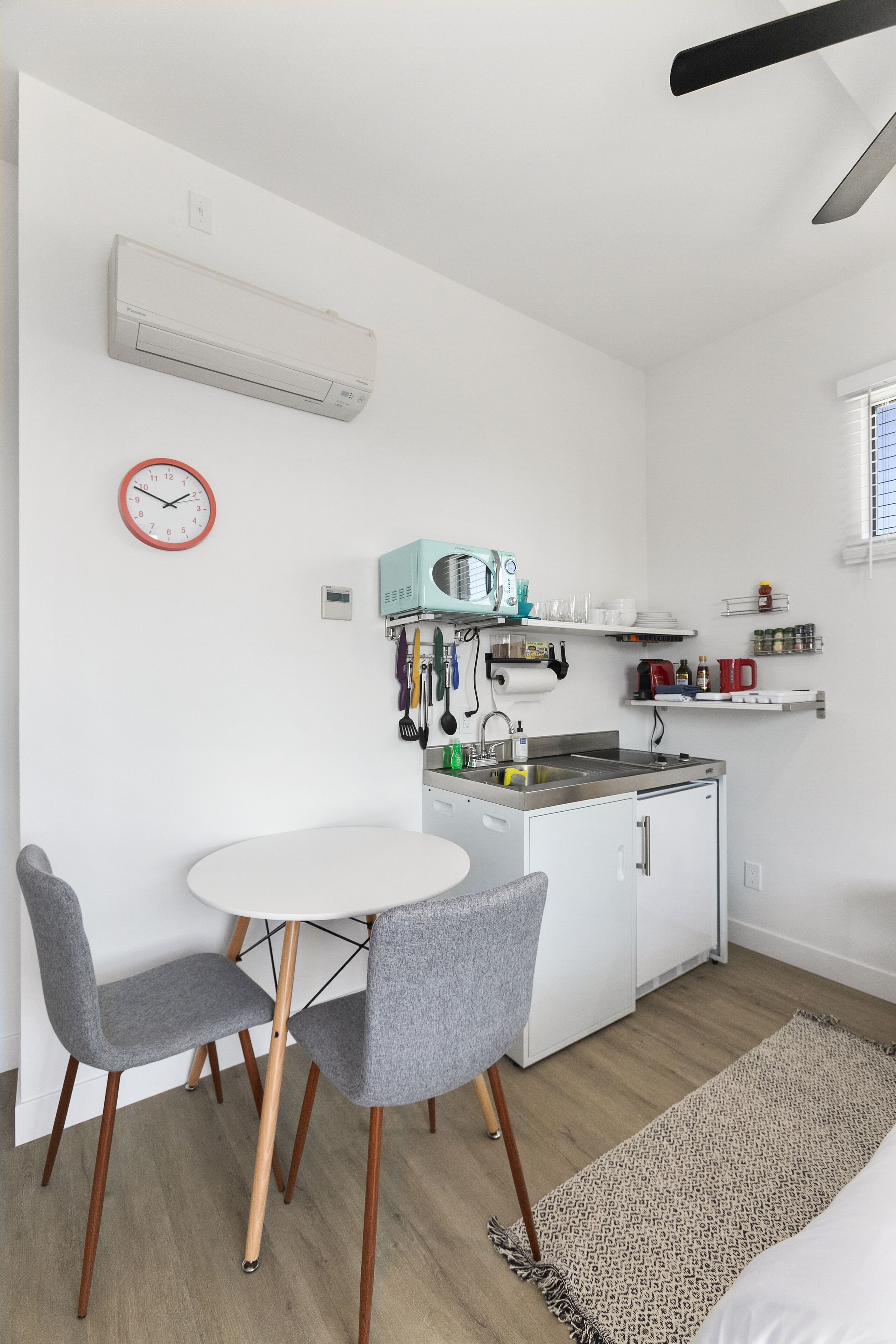 Uma cozinha compacta com geladeira branca, pia pequena e eletrodoméstico verde-água. Há uma área de refeições com uma mesa redonda e duas cadeiras cinzas. Um relógio é visível na parede, e uma janela com persianas está à direita.