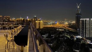 Um bar no terraço com um balcão comprido e cadeiras, com vista para um horizonte urbano com edifícios iluminados e uma autoestrada.