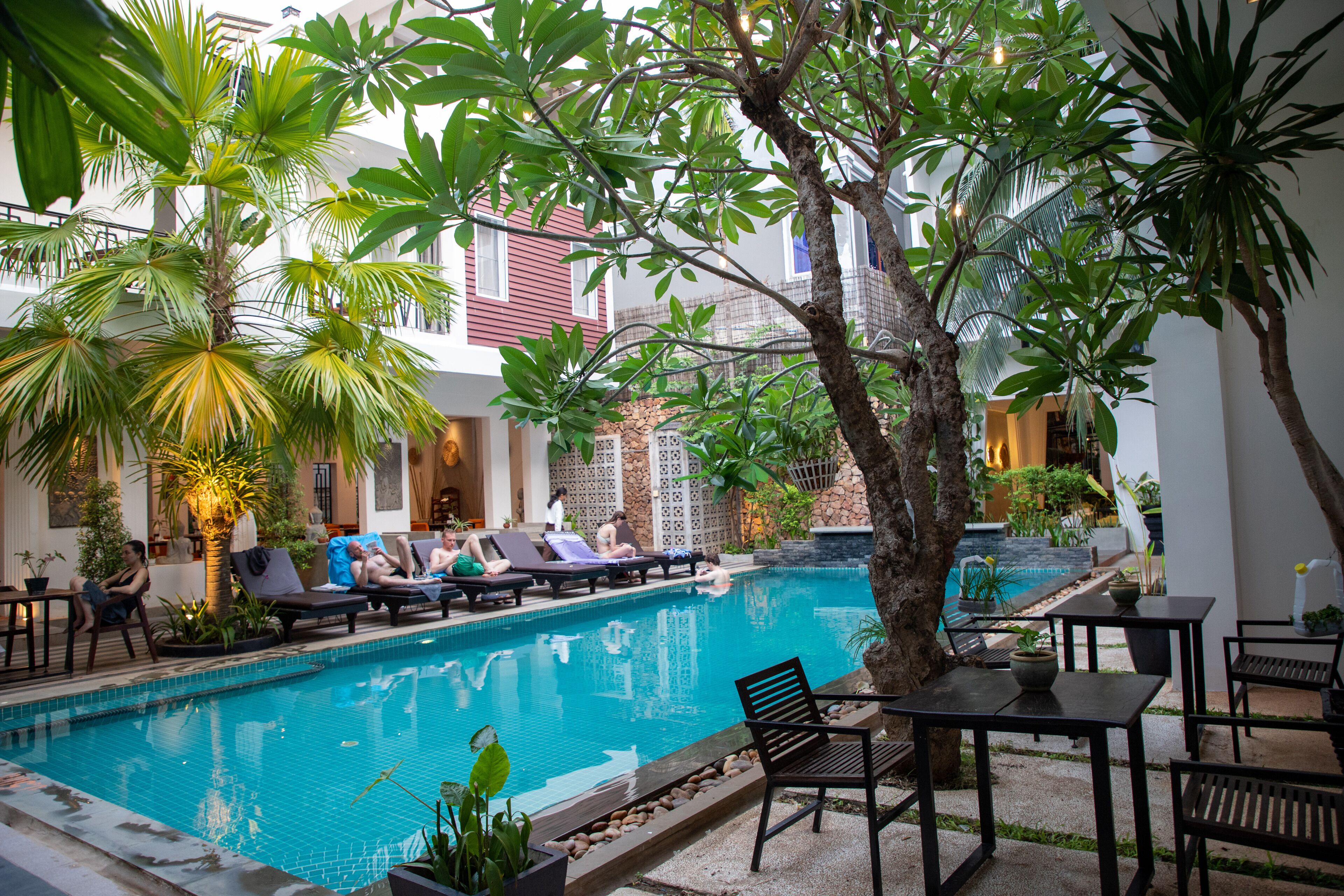 Área de piscina com espreguiçadeiras e mesas, cercada por plantas tropicais e uma piscina cristalina.