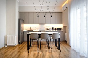 Uma cozinha moderna com bancada de mármore, forno embutido e geladeira. Há três banquetas e uma janela com cortinas leves.