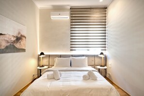 Quarto de hotel moderno com uma cama grande, duas mesas de cabeceira com luminárias, ar condicionado no teto e uma janela ampla com persianas.