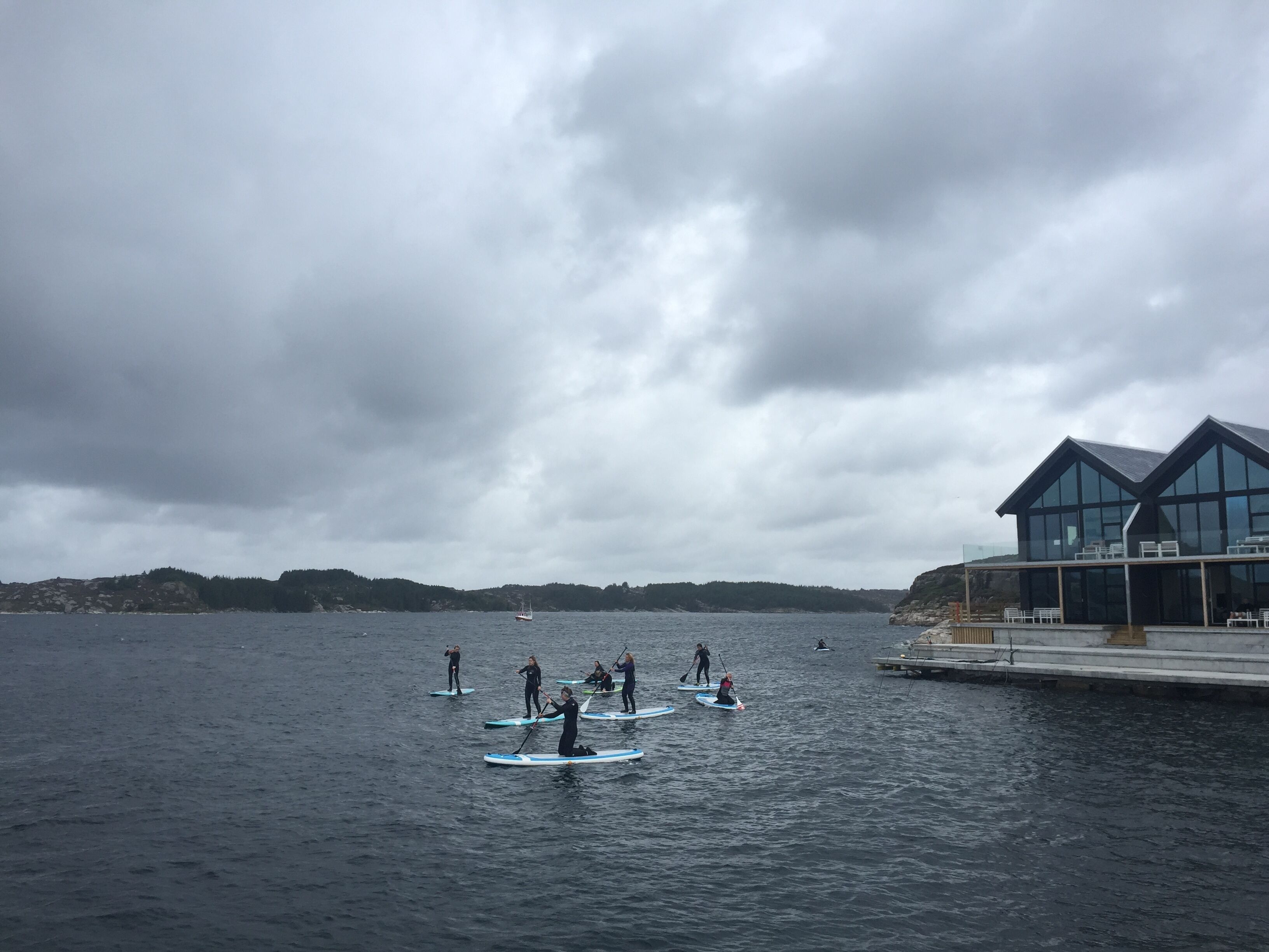 Um edifício à beira-mar com um cais e um grupo de pessoas praticando paddleboarding na água.