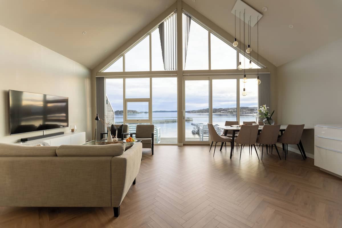Uma sala de estar ampla, com uma televisão grande, um sofá, uma mesa de jantar com cadeiras e vista para o mar através de amplas janelas.