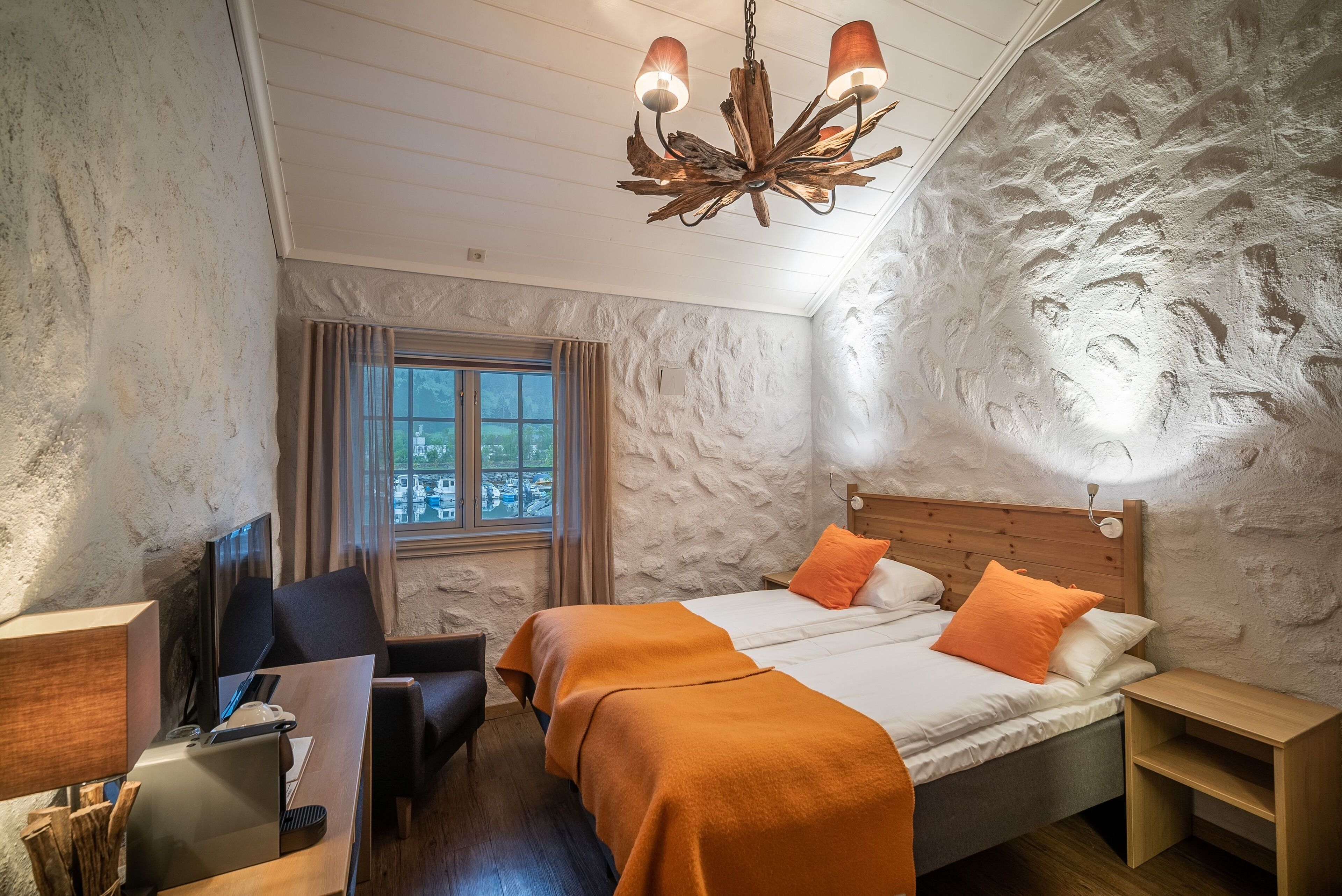 Quarto com parede de pedra rústica, cama de madeira com roupa de cama laranja, escrivaninha com um laptop e janela com cortinas.
