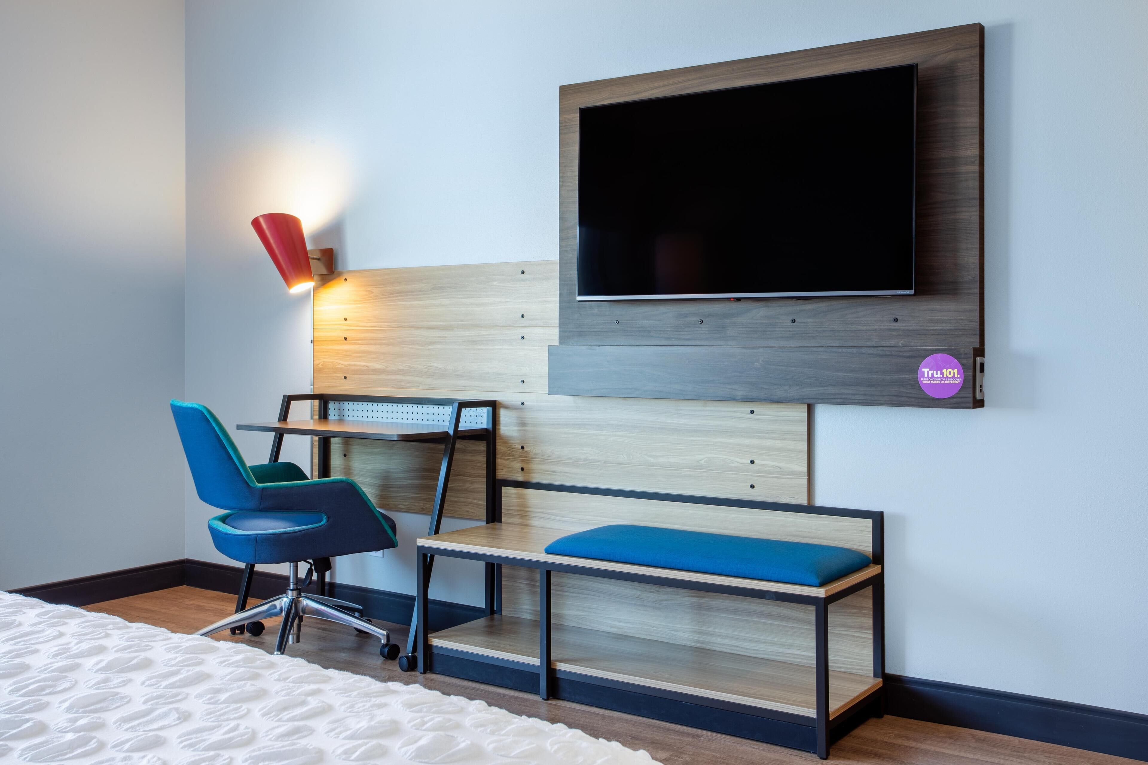 Quarto moderno com televisão fixada numa parede de madeira, uma cadeira azul, um banco e um abajur vermelho.