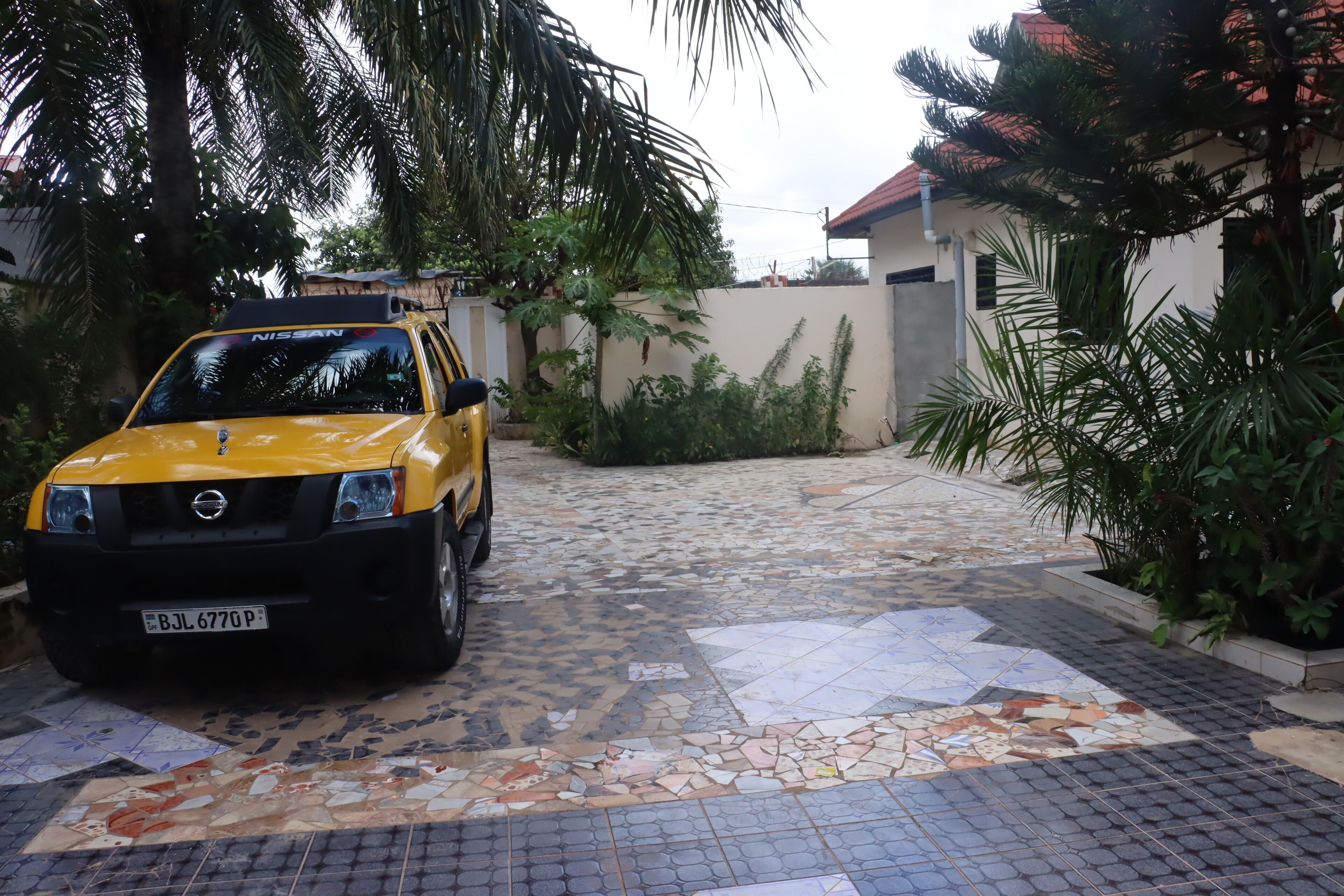 Uma caminhonete Nissan amarela estacionada em uma entrada com piso de mosaico e cercada por vegetação.