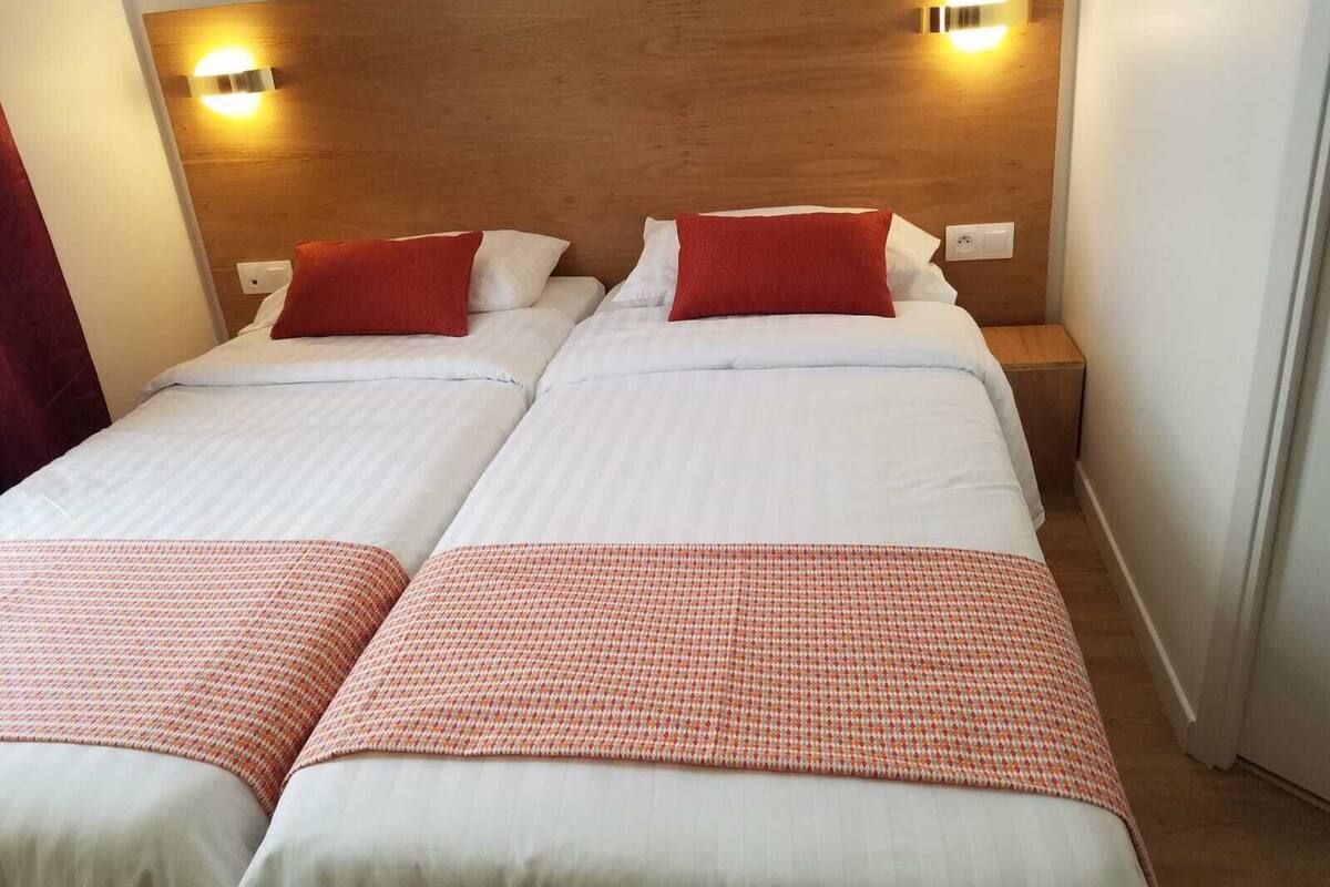 Quarto duplo com duas camas, cada uma com roupa de cama branca e almofadas vermelhas. Duas luminárias fixadas na parede sobre as camas e uma cabeceira de madeira.