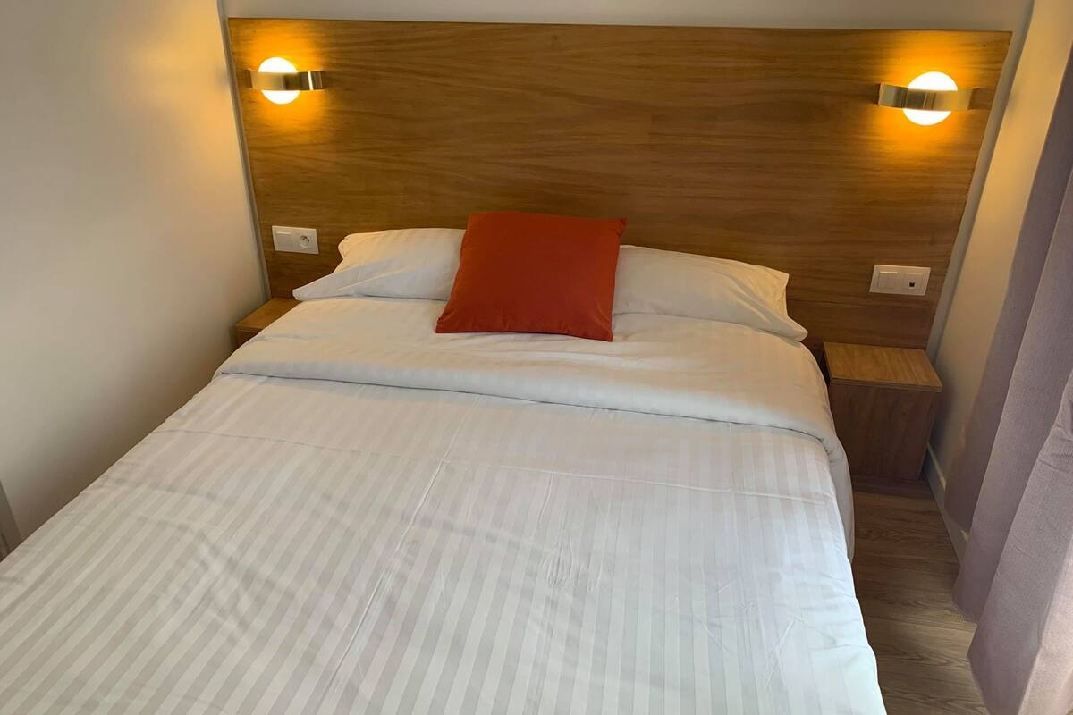 Cama bem arrumada com roupa de cama branca e um único travesseiro vermelho, com duas mesas de cabeceira e mesas laterais de madeira.