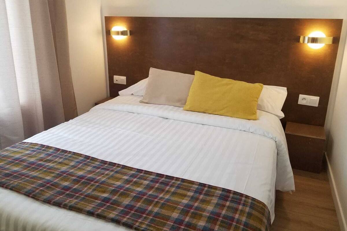 Quarto de hotel com uma cama, duas luminárias de cabeceira, um painel de cabeceira e um cobertor xadrez.
