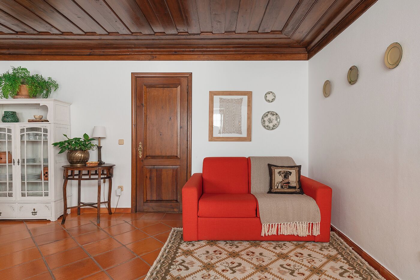 Uma sala de estar com um sofá vermelho, um tapete estampado, uma porta de madeira, um armário com portas de vidro e uma mesinha com abajur.