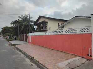 Uma rua residencial com um muro vermelho, calçada pavimentada e uma casa com varanda.
