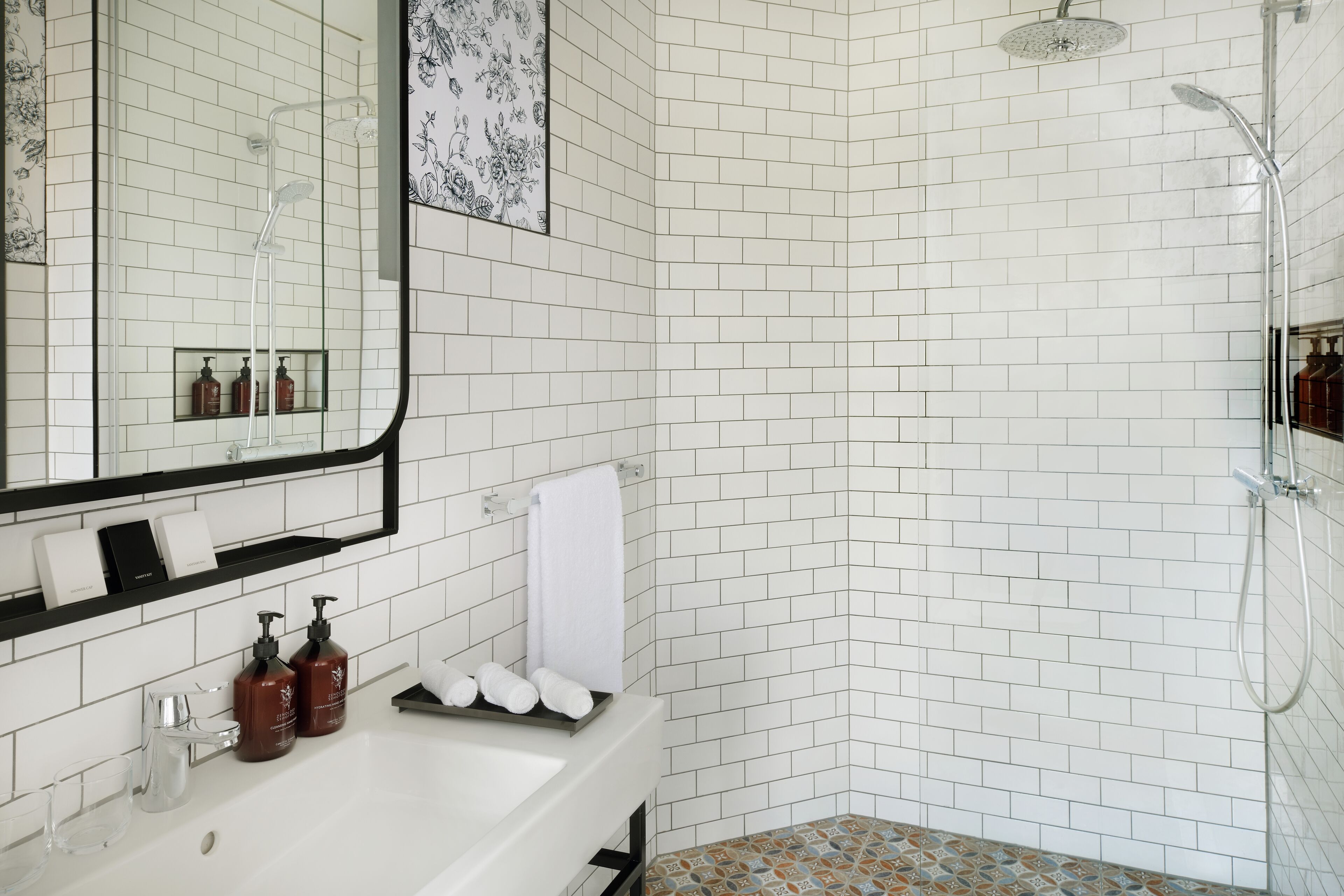 Um banheiro moderno com azulejos brancos tipo metrô, box de vidro, pia branca com prateleira preta e piso de azulejos estampados.