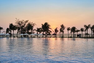Vista da piscina ao pôr do sol, com palmeiras e guarda-s óis.