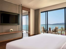 Quarto de hotel com uma cama grande, televisão de tela plana na parede, varanda com mesa e cadeiras, e vista para o mar.