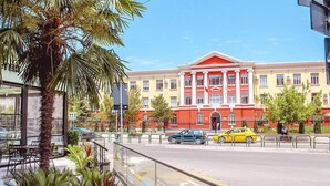 Um edifício com fachada vermelha e colunas brancas, situado numa esquina com palmeiras e um café com mesas na calçada.