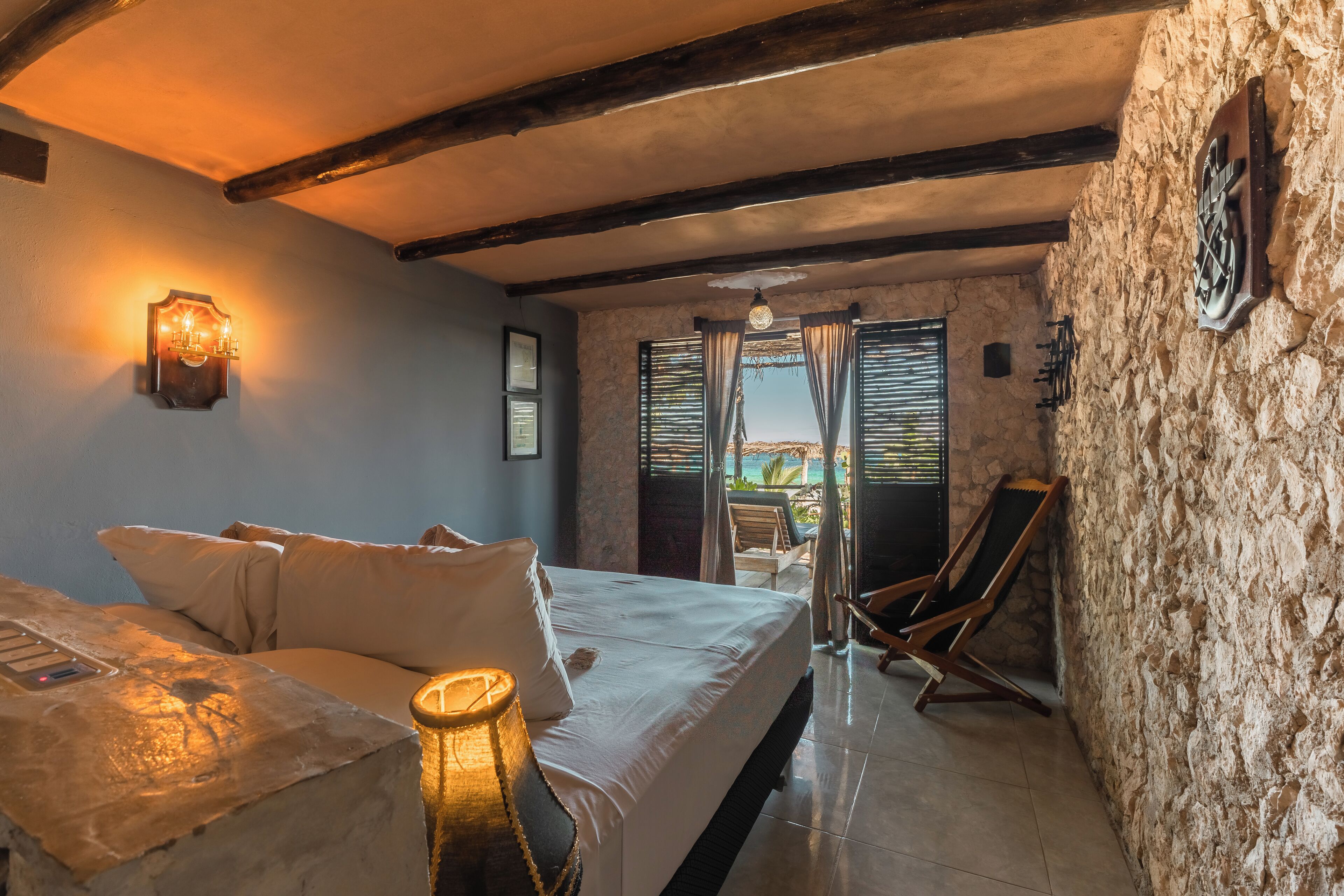 Quarto com cama, mesa de cabeceira, luminária na parede, janela com cortinas e parede de pedra.