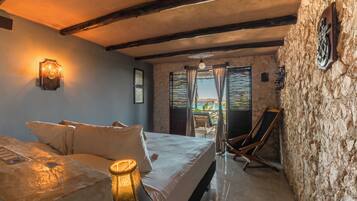 Quarto com cama, mesa de cabeceira, luminária na parede, janela com cortinas e parede de pedra.
