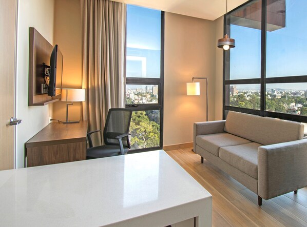 Quarto de hotel com escrivaninha, cadeira, sofá, TV fixada na parede, luminária e amplas janelas com vista para a cidade.