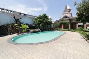 Piscina com um calçadão azulejado, rodeada por vegetação e um edifício com cúpula e torre.