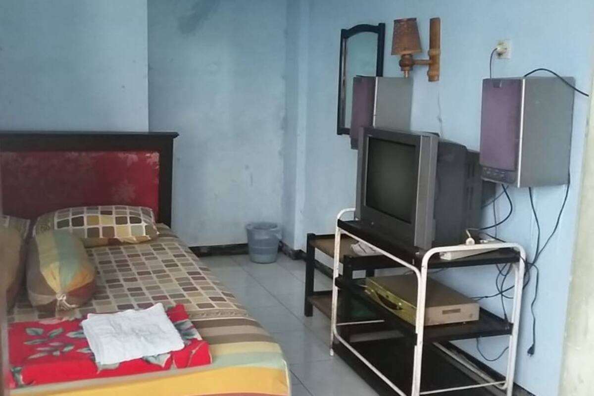 Quarto pequeno e simples, com uma cama de solteiro, televisão sobre um suporte e uma lixeira.