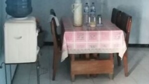 Uma pequena sala de jantar com uma mesa posta para dois, um bebedouro, uma geladeira e uma televisão na parede.