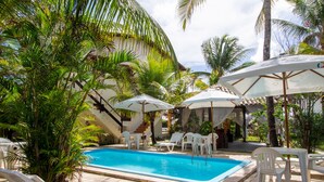 Uma área de piscina com cadeiras de lounge brancas e guarda-sóis, cercada por palmeiras e um deck revestido de azulejos.