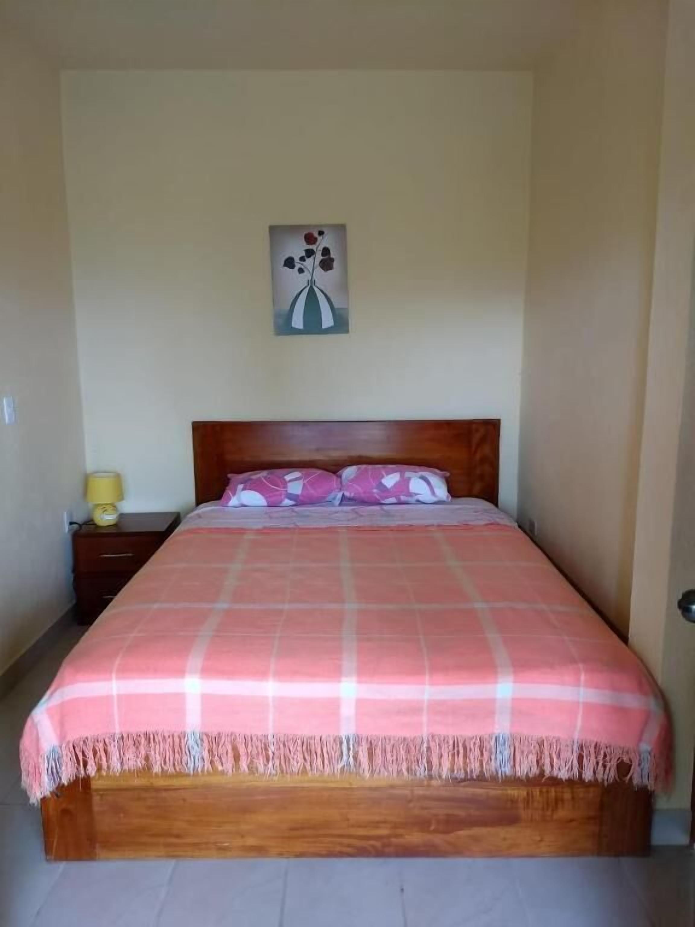 Um quarto com cama de madeira, colcha xadrez rosa e branca e uma pequena mesa de cabeceira com abajur.