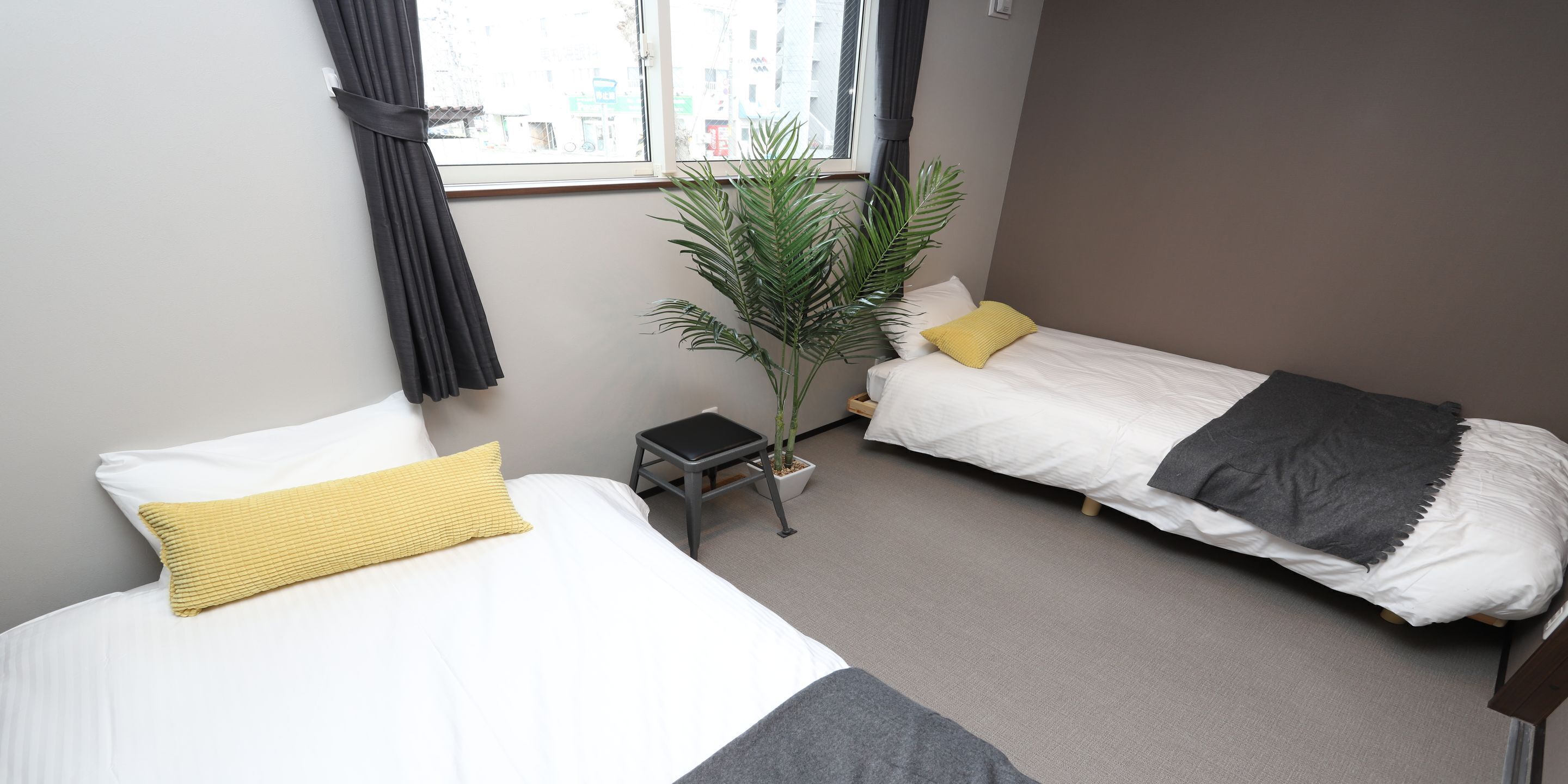 Quarto de hotel com duas camas, uma mesinha, um vaso de planta e uma janela com cortinas.