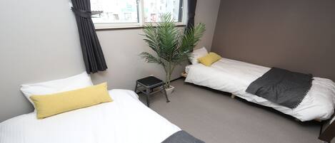 Quarto de hotel com duas camas, uma mesinha, um vaso de planta e uma janela com cortinas.