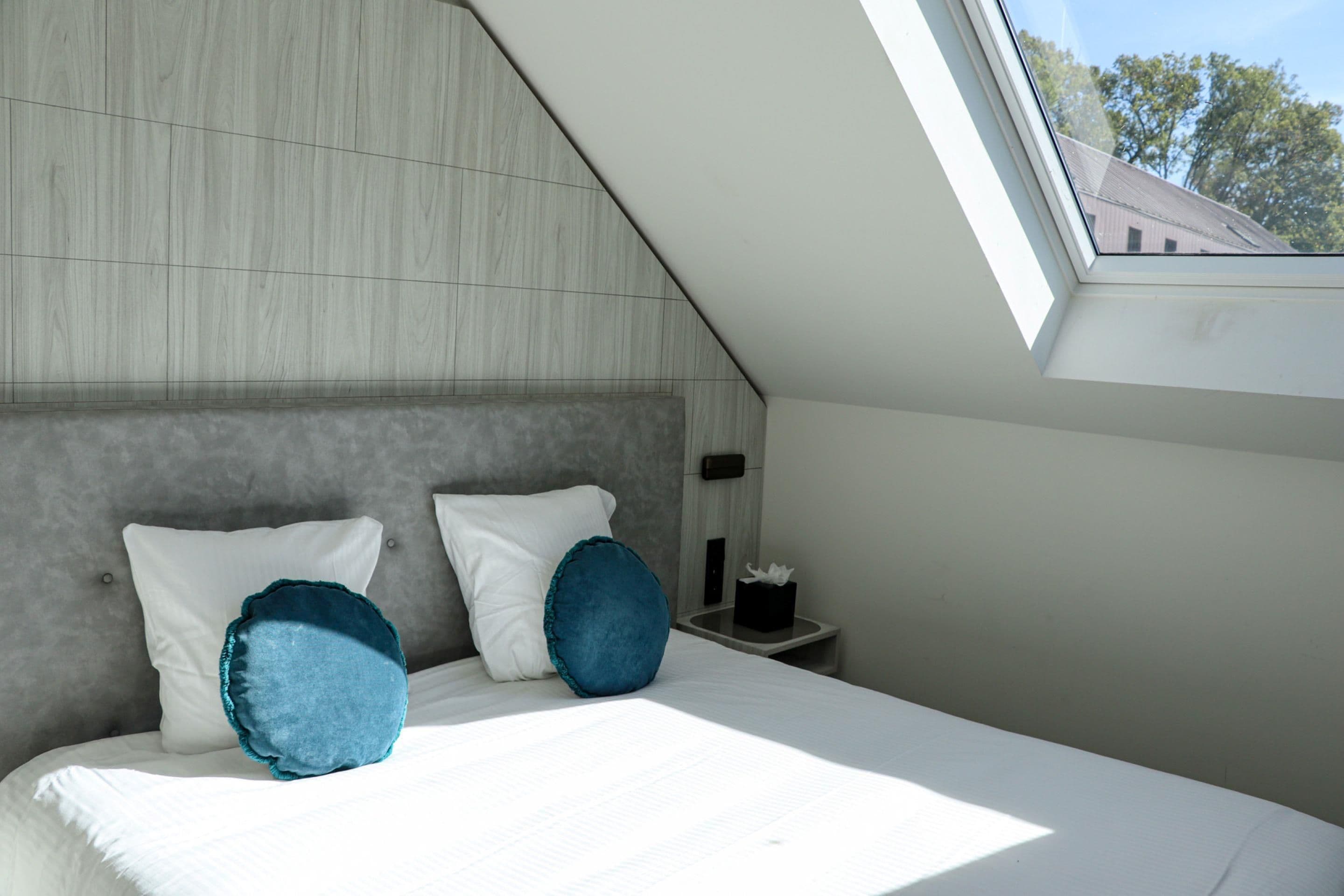 Quarto com teto inclinado, cama com roupa de cama branca, duas almofadas, mesa de cabeceira com abajur e janela com vista para prédios.