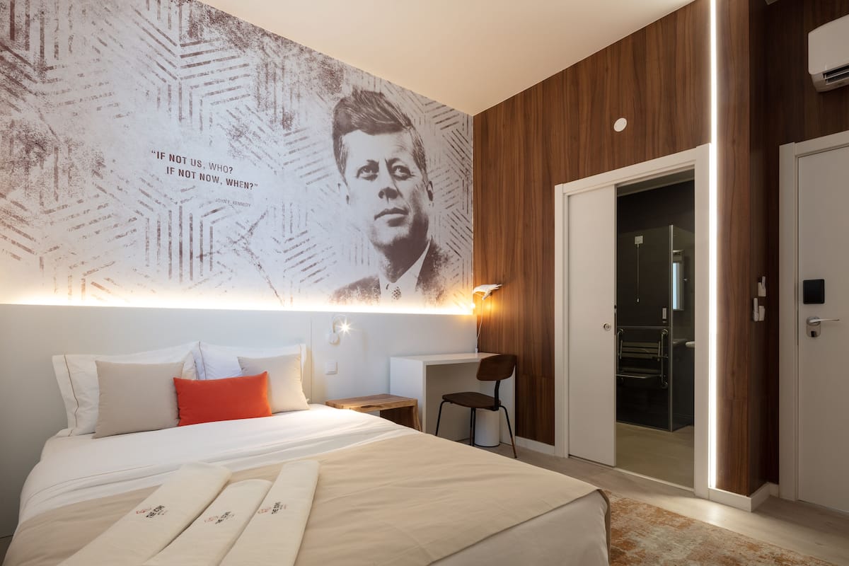 Quarto de hotel moderno com uma cama grande, uma escrivaninha e uma frase decorativa na parede.