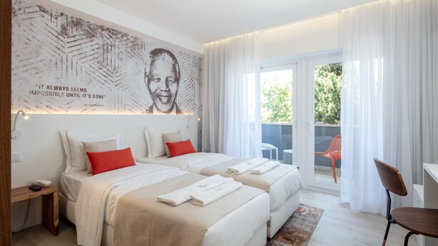 Quarto de hotel com duas camas, uma escrivaninha, uma cadeira e varanda com vista.