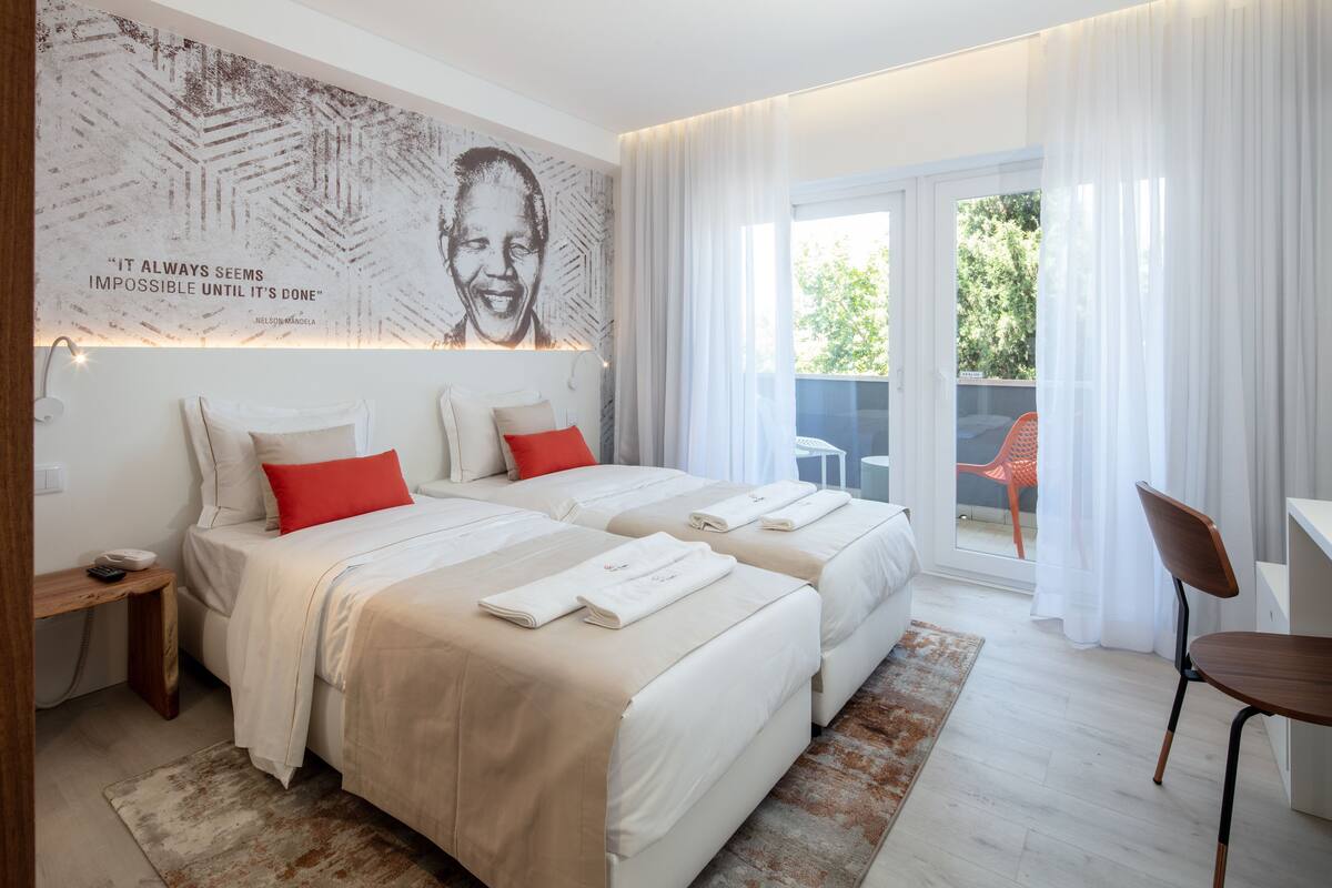 Quarto de hotel com duas camas, uma escrivaninha, uma cadeira e varanda com vista.