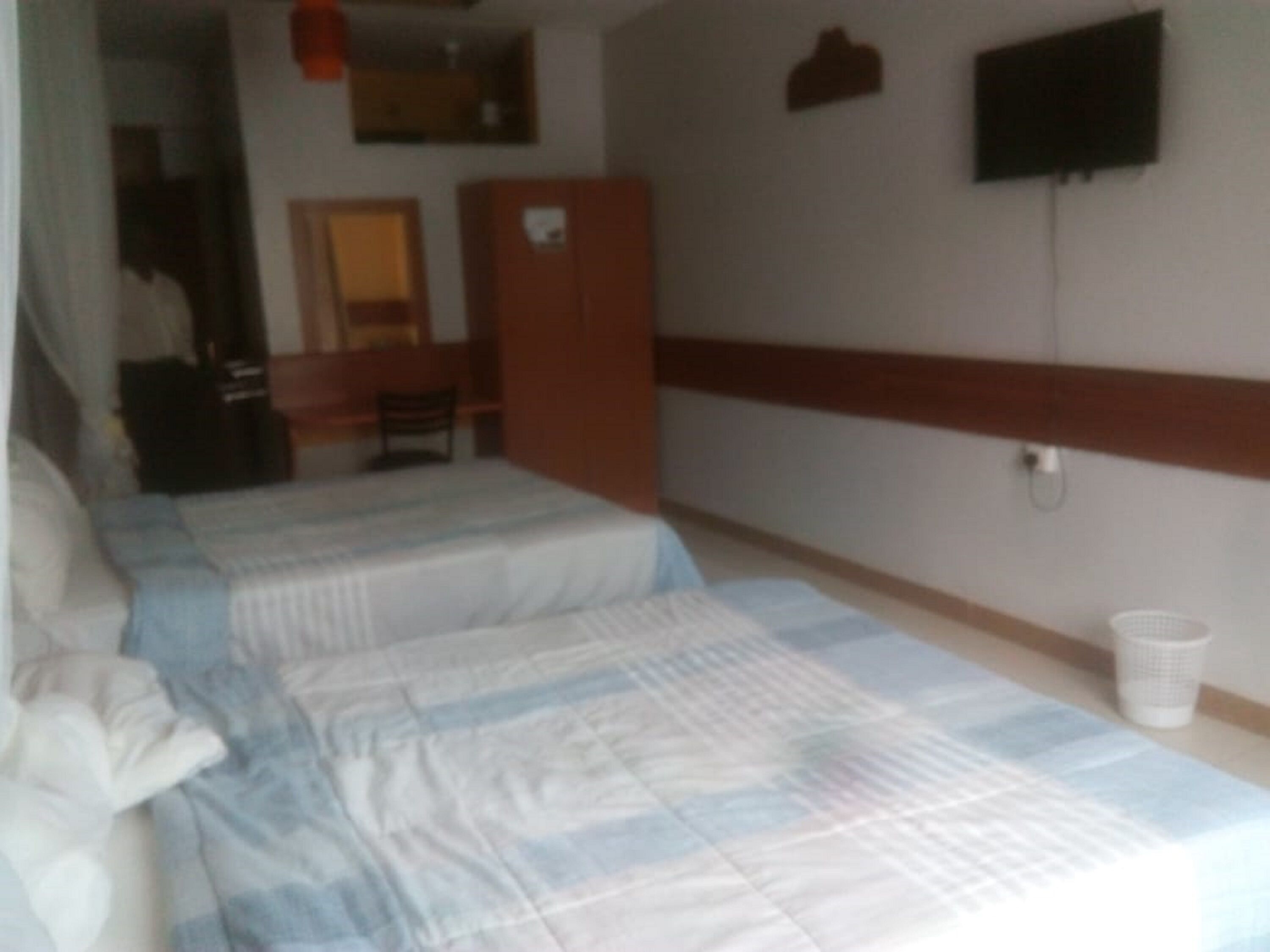 Quarto de hotel com duas camas, uma escrivaninha, uma cadeira, um guarda-roupa e uma televisão na parede.