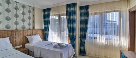 Quarto de hotel com duas camas, cabeceira de madeira, uma janela grande com cortinas leves e papel de parede estampado.