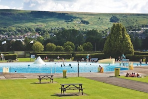 Piscina com uma fonte, rodeada por uma área gramada com mesas de piquenique e vista para uma cidade ao fundo.