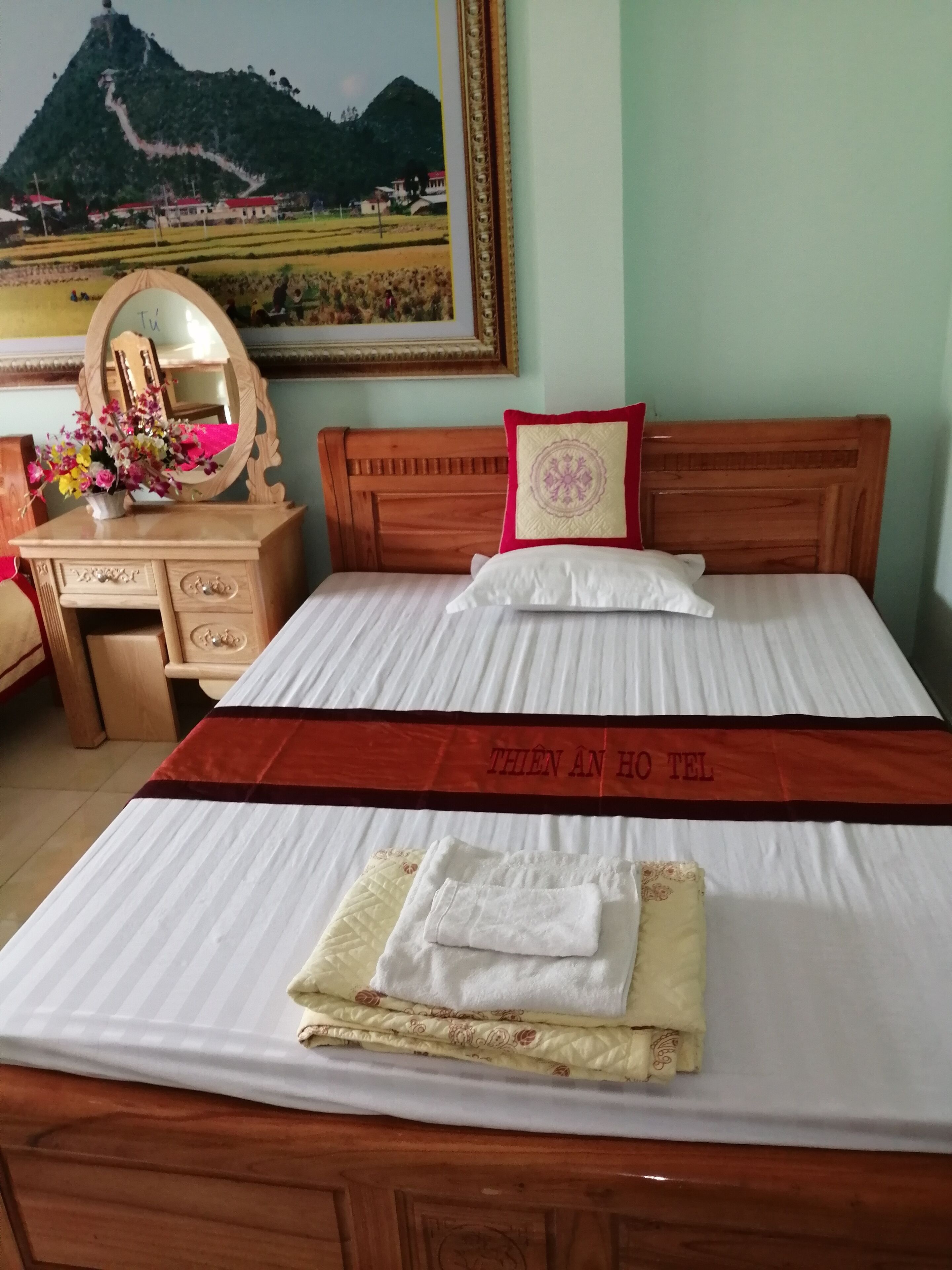 Uma cama bem arrumada com colcha vermelha e branca, cabeceira de madeira e travesseiro com padrão floral. Um criado-mudo de madeira com espelho e um vaso de flores. Uma moldura com uma paisagem montanhosa na parede.