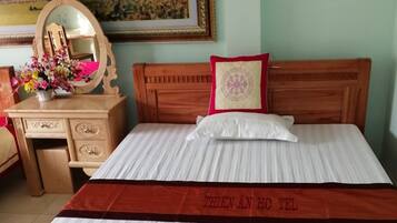 Uma cama bem arrumada com colcha vermelha e branca, cabeceira de madeira e travesseiro com padrão floral. Um criado-mudo de madeira com espelho e um vaso de flores. Uma moldura com uma paisagem montanhosa na parede.