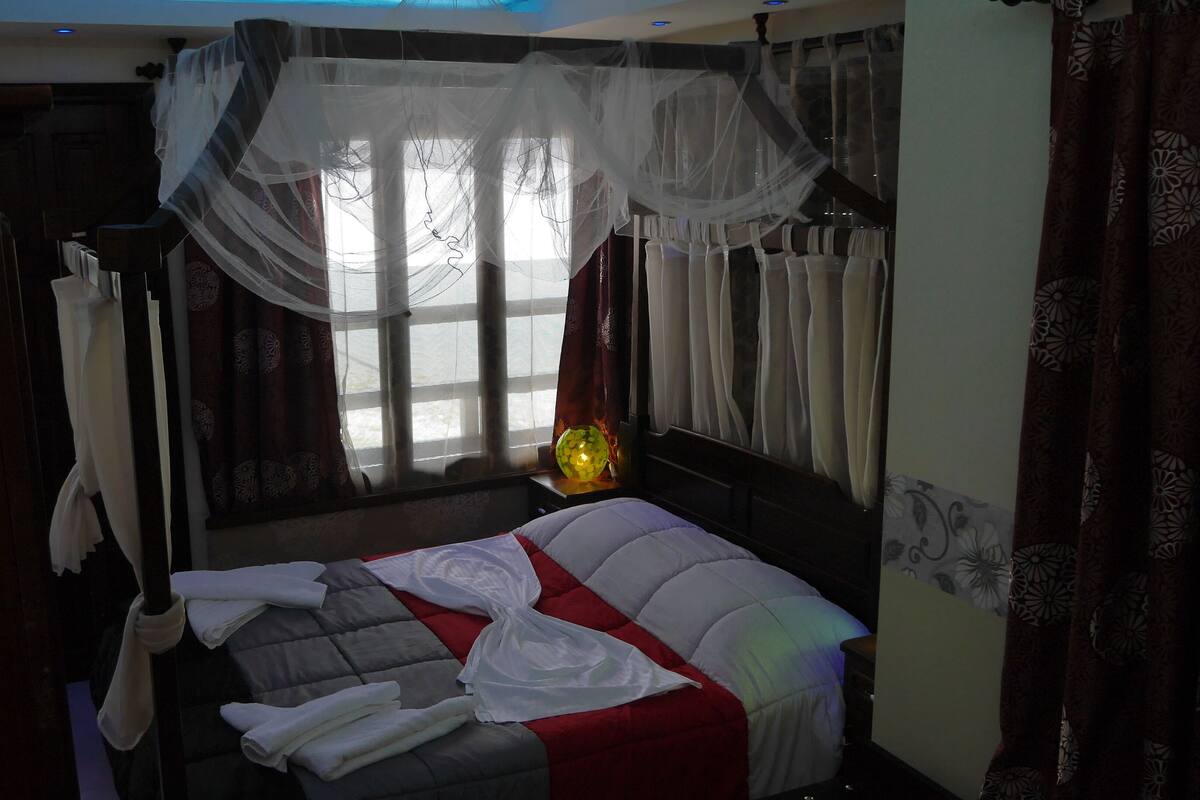 Quarto de hotel com cama, mesa de cabeceira, abajur e janela com cortinas.
