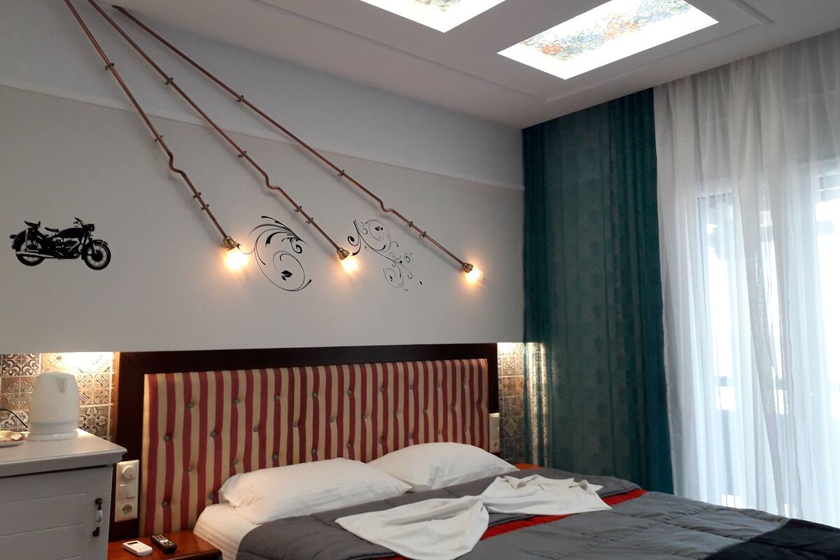 Quarto de hotel com cama, mesa de cabeceira, luminária na parede e janela com cortinas.