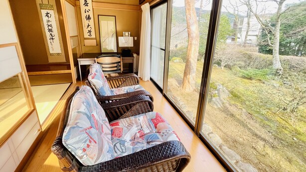 Um quarto japonês tradicional com piso de tatame, uma mesa baixa, um banco acolchoado e uma grande janela com vista para um jardim.
