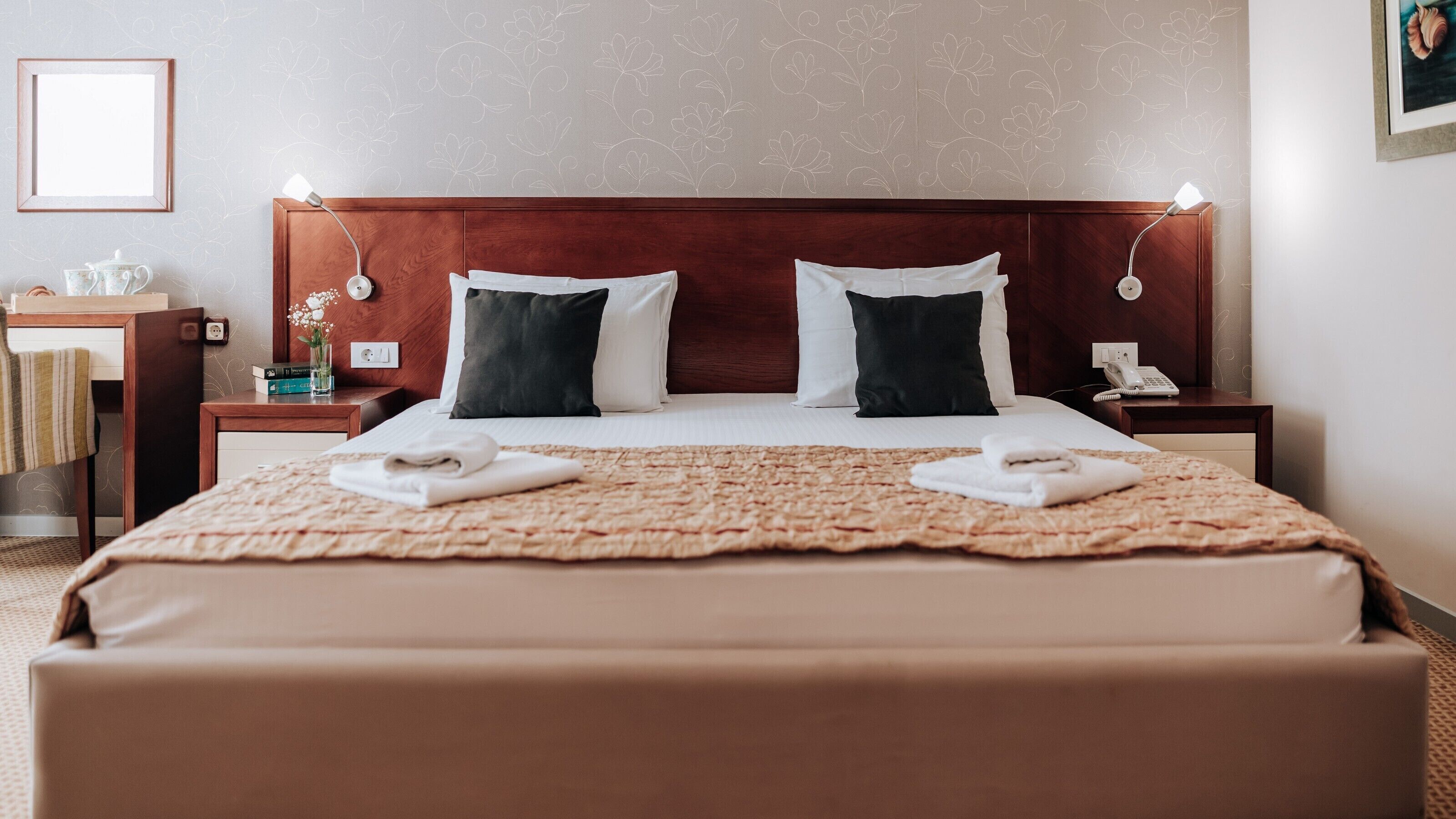 Cama bem arrumada com cobertura marrom, duas luminárias de cabeceira, mesa de cabeceira com telefone e parede com padrão floral.