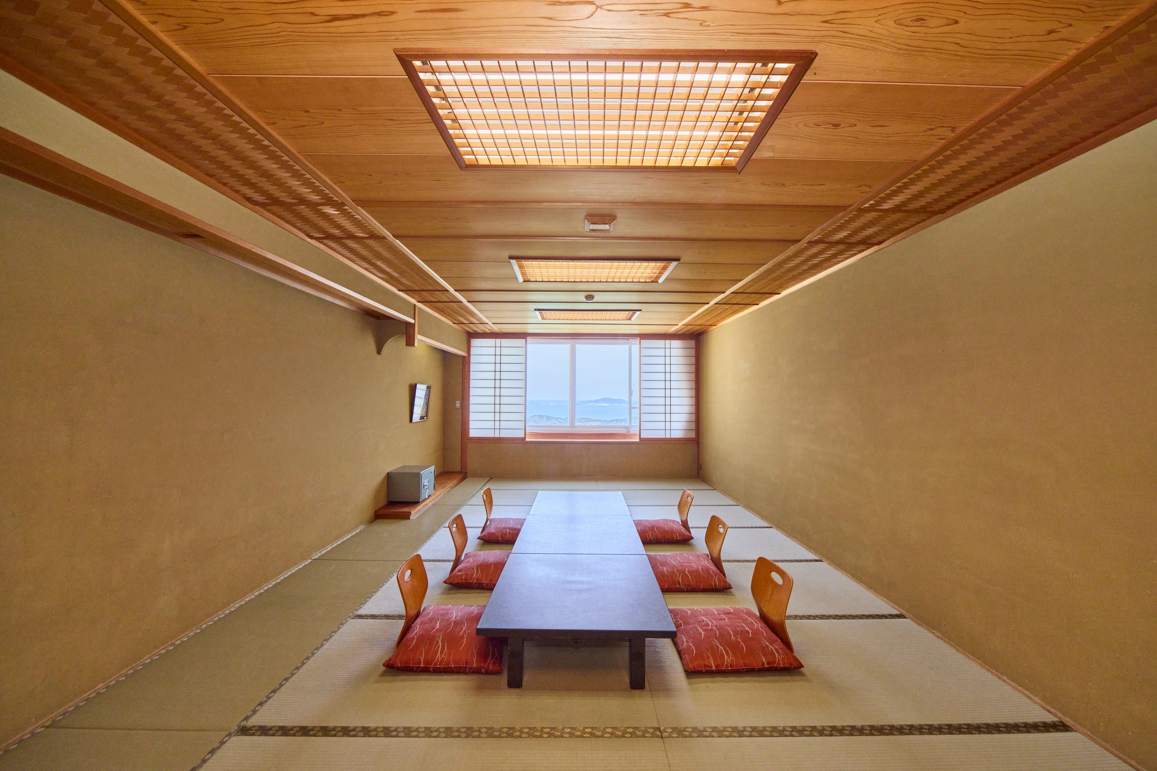 Um quarto tradicional em tatame, com uma mesinha baixa e almofadas, vigas de madeira no teto e vista através de um painel shoji.