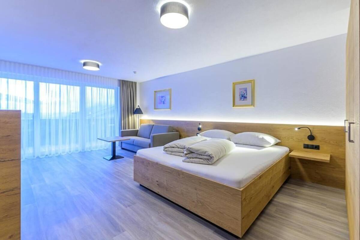 Quarto de hotel moderno com uma cama de madeira, uma escrivaninha e uma cadeira. Há duas molduras com quadros na parede e uma janela com cortinas.